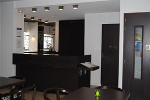 Uma área de recepção com um balcão escuro, um mostrador e uma porta com uma placa. Há cadeiras e uma mesa em primeiro plano.