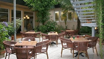 Uma área de café ao ar livre com mesas e cadeiras de vime, cercada por vegetação e plantas em vasos.