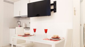 Cozinha moderna com armários brancos, uma TV de tela plana na parede e uma área de jantar com mesa de vidro e cadeiras brancas.