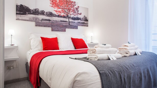 Cama bem arrumada com roupa de cama vermelha e branca, um cobertor cinza e um conjunto de toalhas ao lado. Há duas mesas de cabeceira com abajures, e um quadro grande com uma imagem de uma árvore na parede.