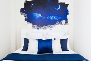 Uma cama com roupa de cama branca e azul, um mural da noite no muro, e uma cabeceira branca.
