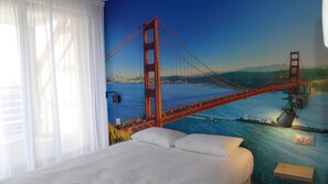 Quarto de hotel com um grande mural da Ponte Golden Gate, uma cama com roupa de cama branca e uma mesa de cabeceira com abajur.