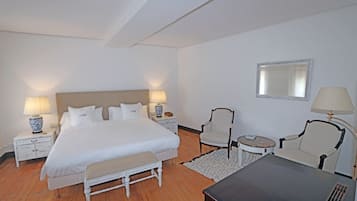 Quarto de hotel com uma cama grande, duas mesas de cabeceira com abajures, uma cadeira, uma pequena mesa redonda e um banco.