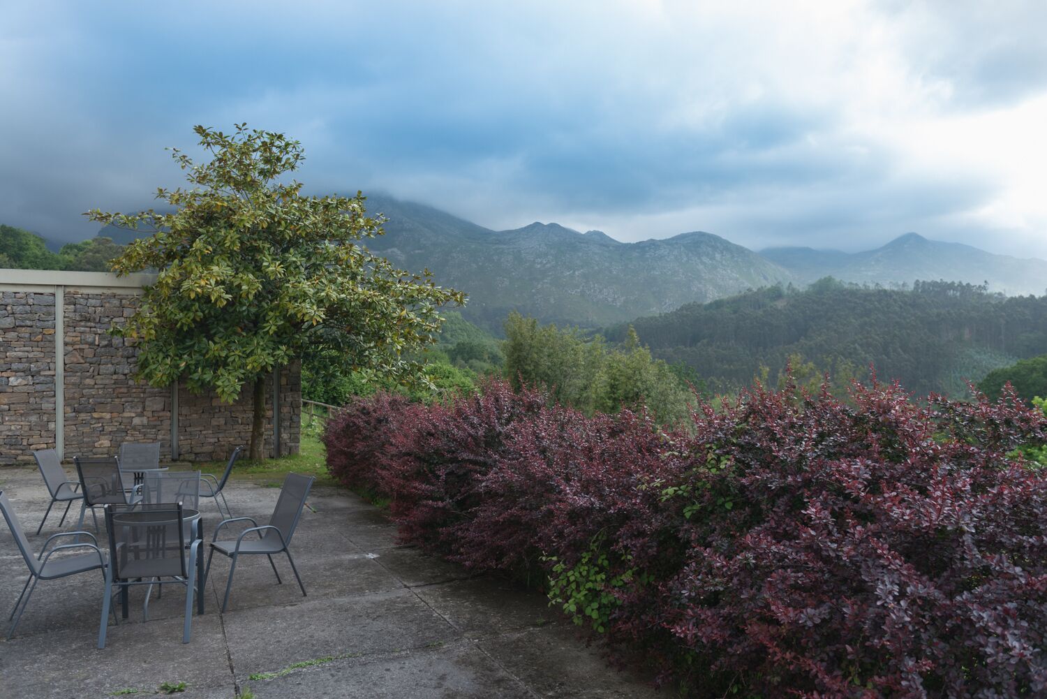 Um pátio com cadeiras e uma mesa, rodeado por um muro de pedra e uma árvore, com vista para uma paisagem montanhosa.