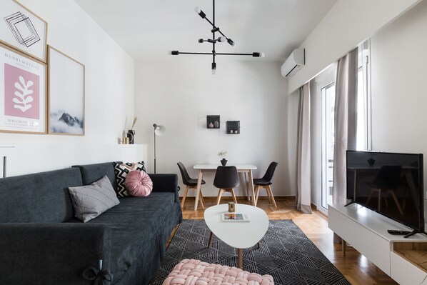 Uma sala de estar moderna com um sofá cinza escuro, uma mesa de centro branca e um banquinho rosa. Há uma área de jantar com uma mesa branca e cadeiras pretas, e uma televisão de tela plana grande fixada em um móvel branco.