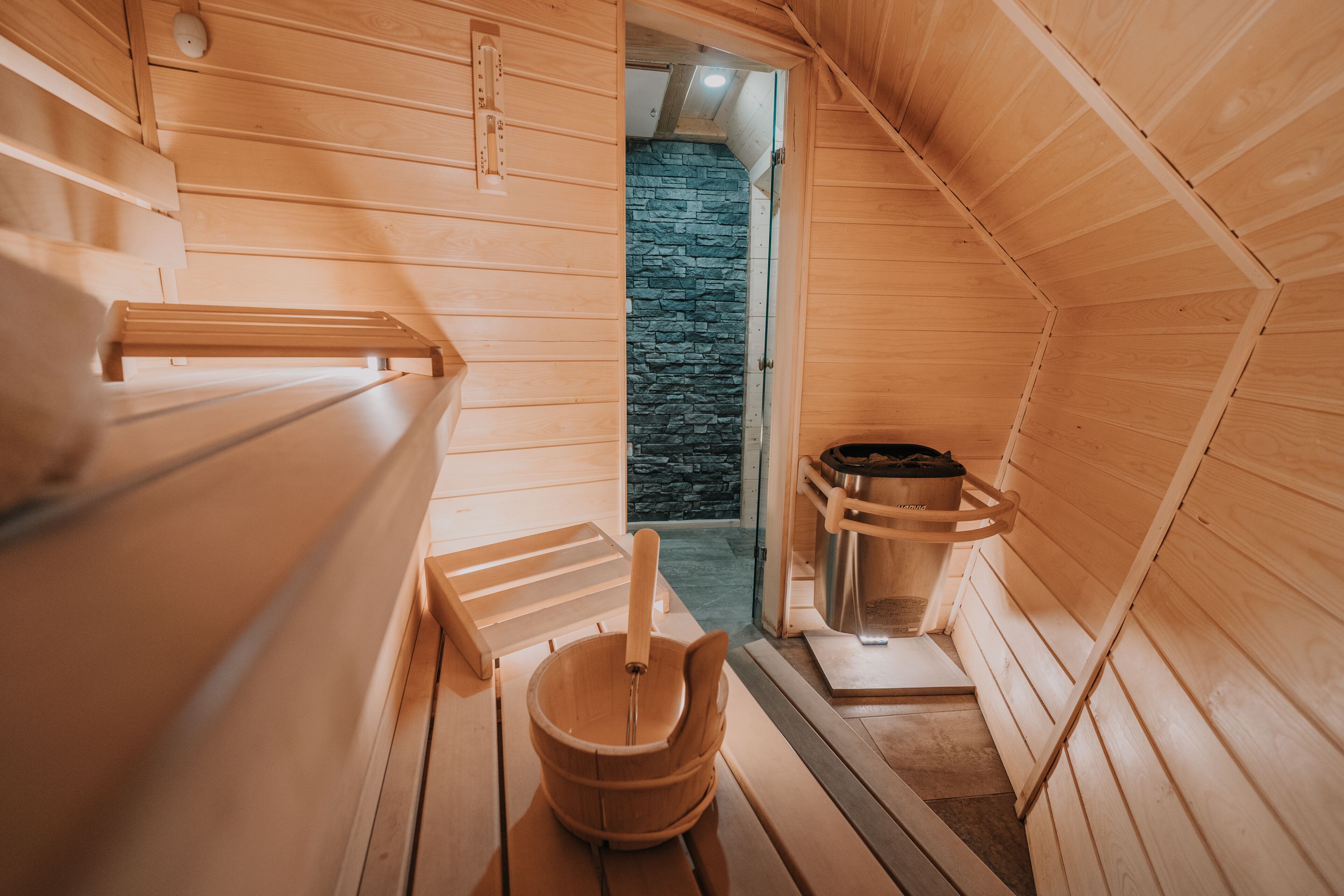 Uma sala de sauna com paredes de madeira, uma parede de pedra ao fundo, um banco de madeira, um balde de madeira e um recipiente de metal.