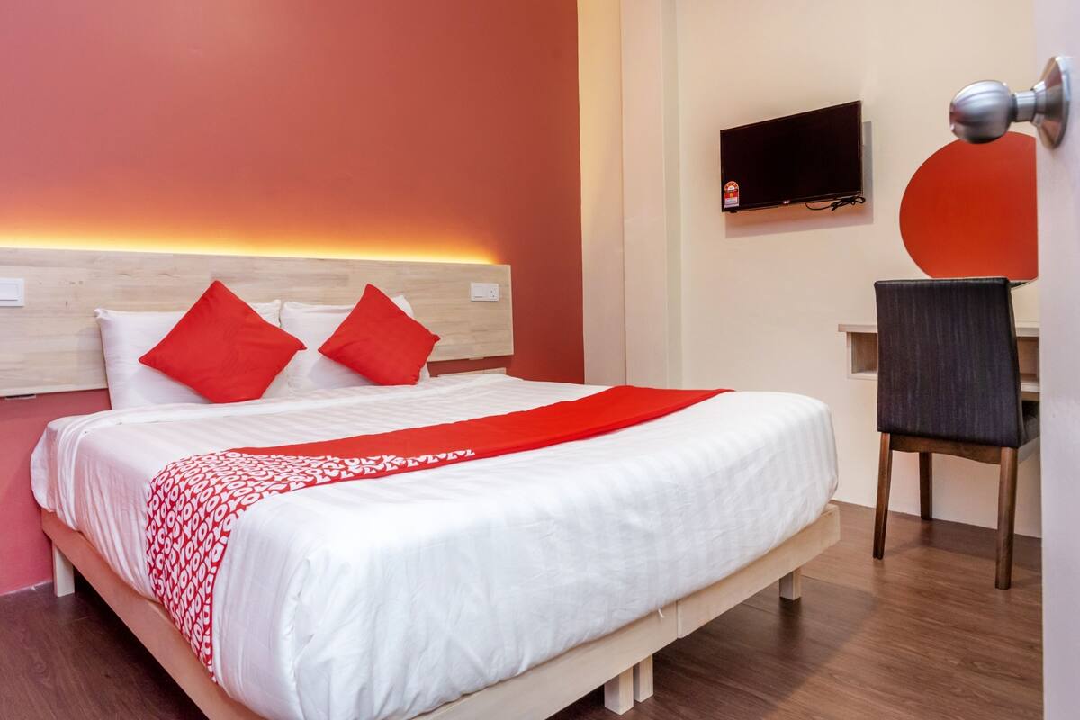 Quarto de hotel moderno com uma cama grande, almofadas vermelhas, manta vermelha, uma escrivaninha com cadeira e uma TV de tela plana fixada na parede.