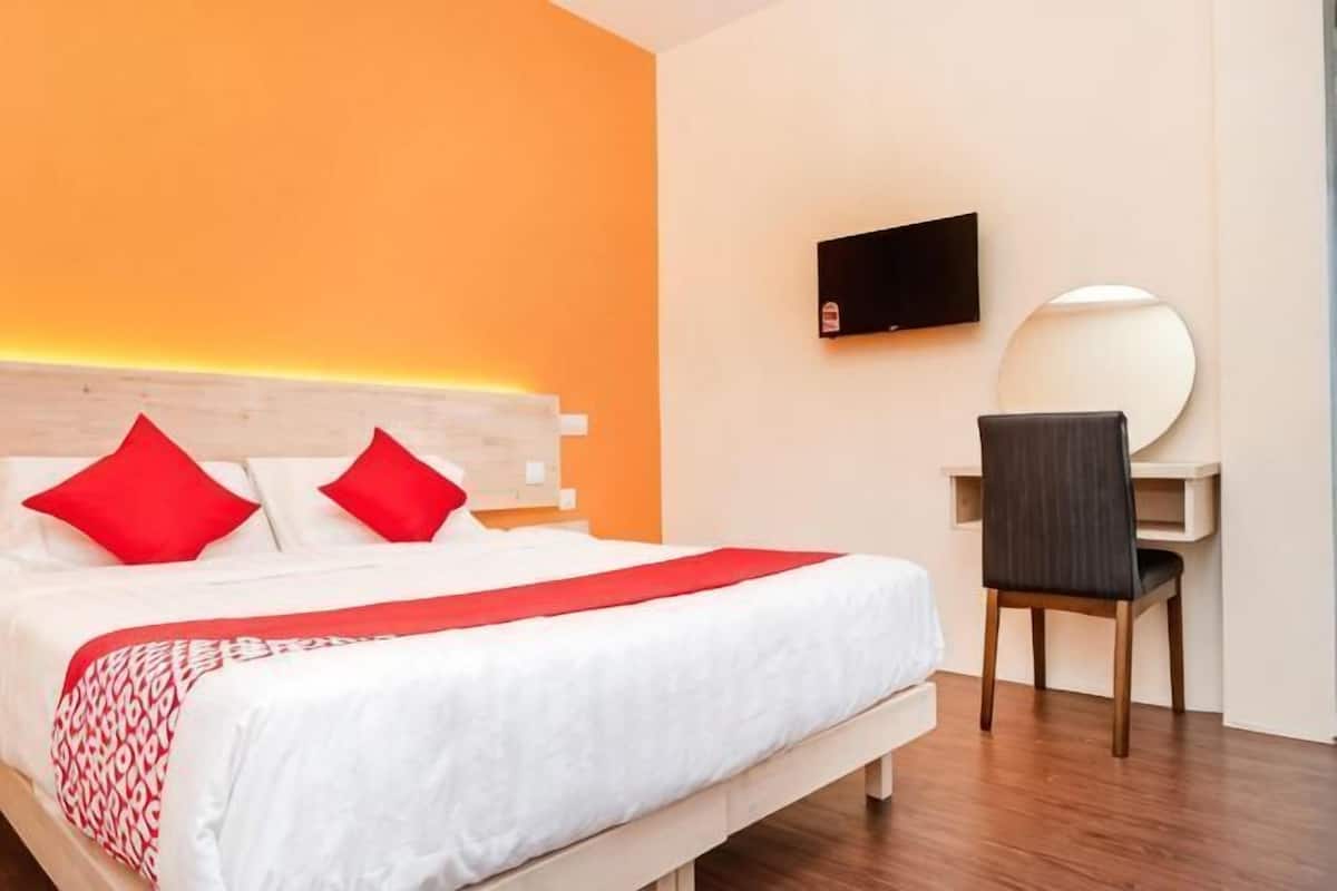 Quarto de hotel com cama, escrivaninha, cadeira, televisão e luminária na parede.