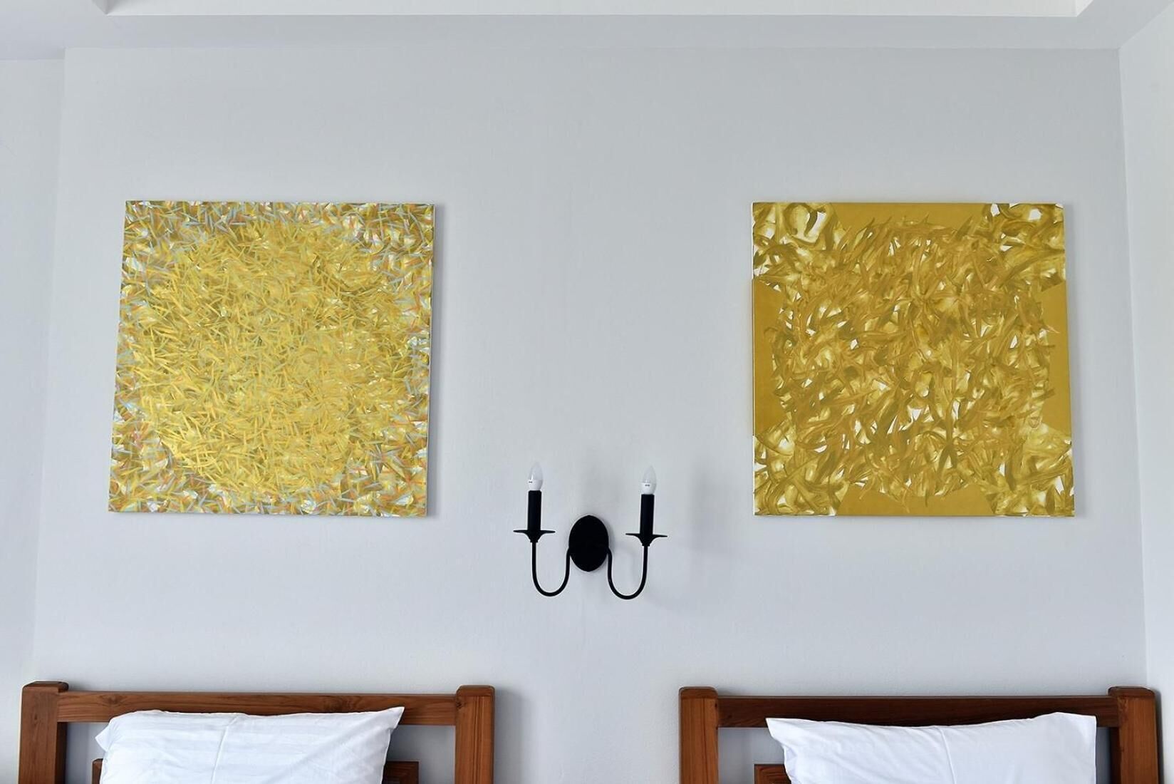 Quarto com duas camas, cada uma com cabeceiras de madeira e roupa de cama branca. Duas arandelas fixadas na parede com abajures pretos e lâmpadas brancas.
