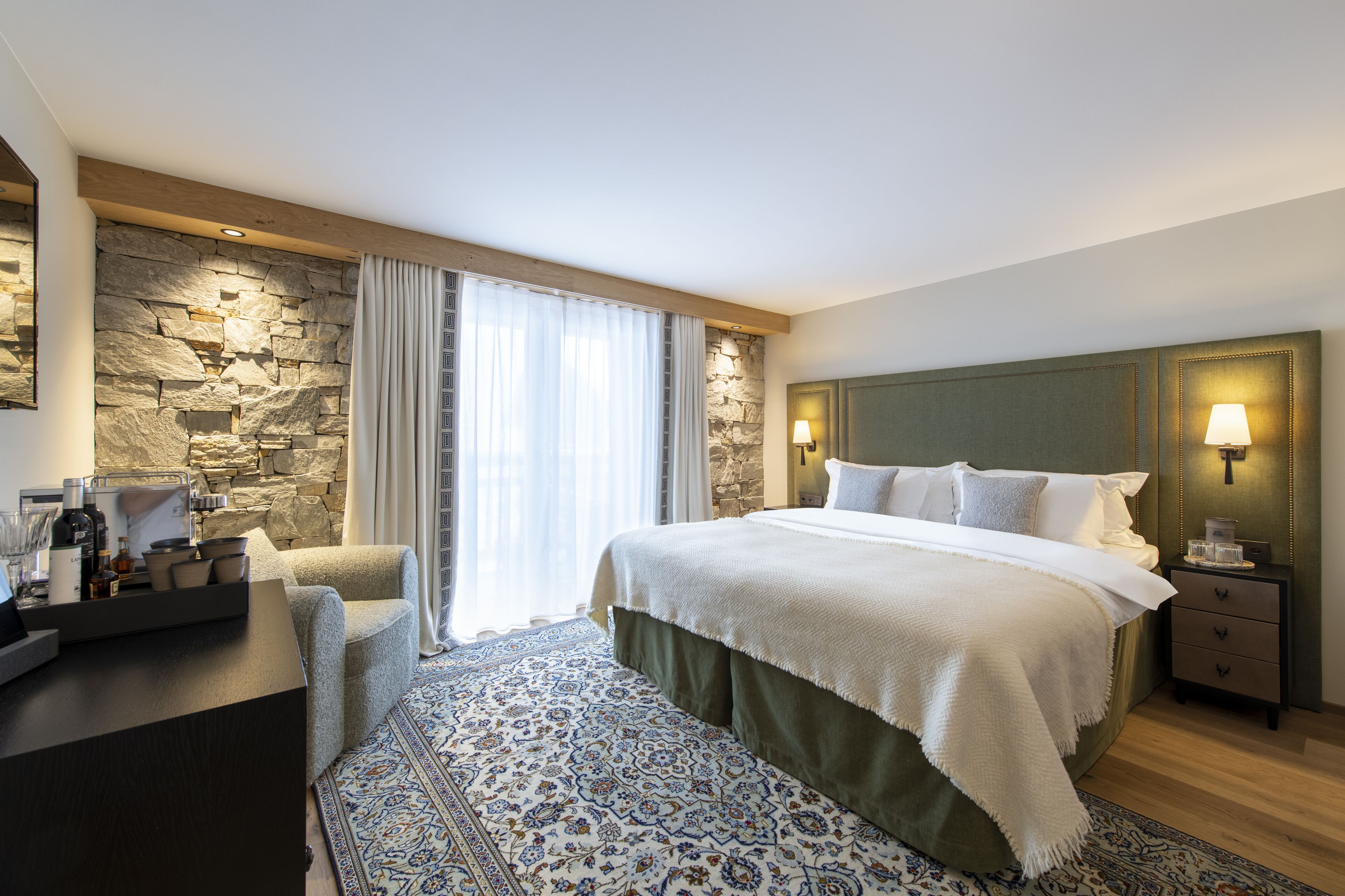 Quarto de hotel com parede de destaque em pedra, uma cama grande com cabeceira verde, uma mesa de trabalho com cadeira e uma mesa de cabeceira com abajur.
