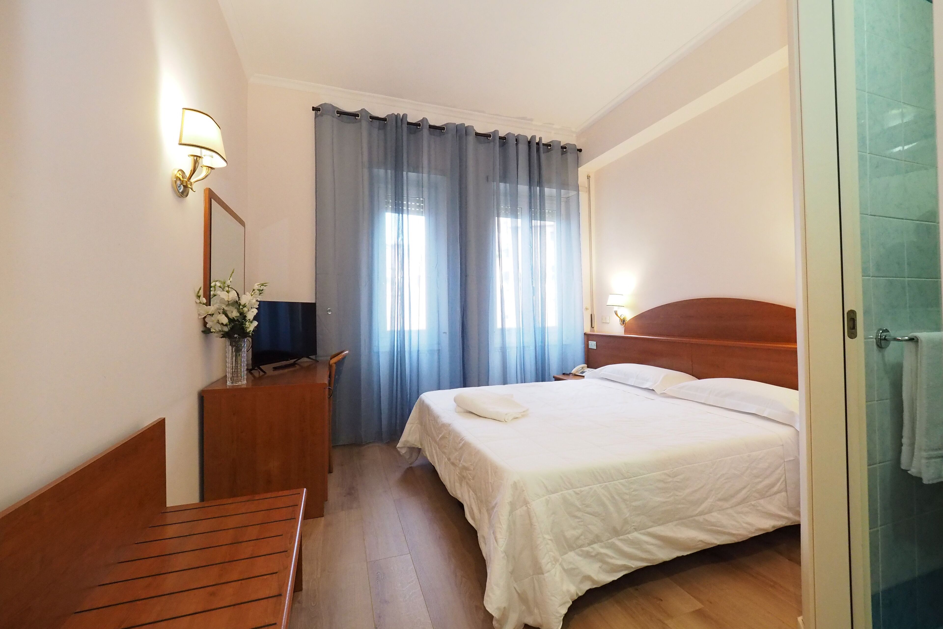 Quarto de hotel com um banco de madeira, mesa de cabeceira com um vaso de flores, cama com roupa de cama branca e box de chuveiro em vidro.