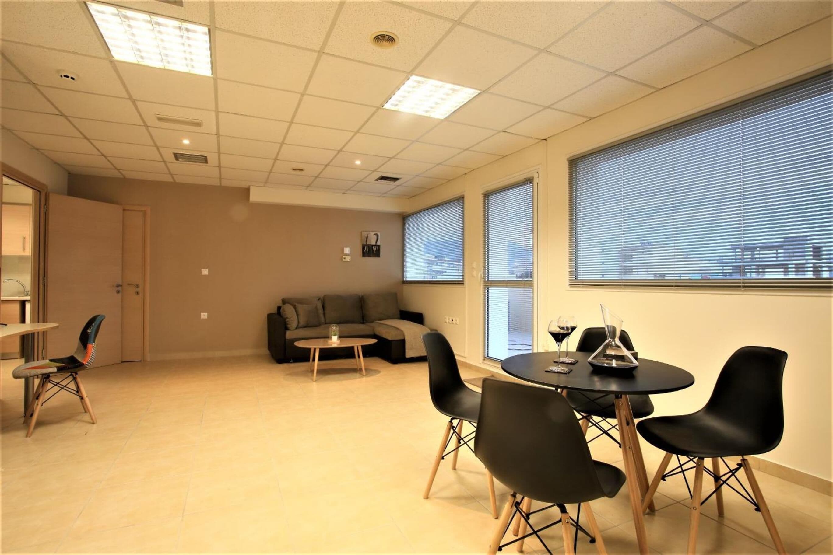 Quarto moderno com mesa redonda, cadeiras pretas, um sofá e janelas amplas com persianas.