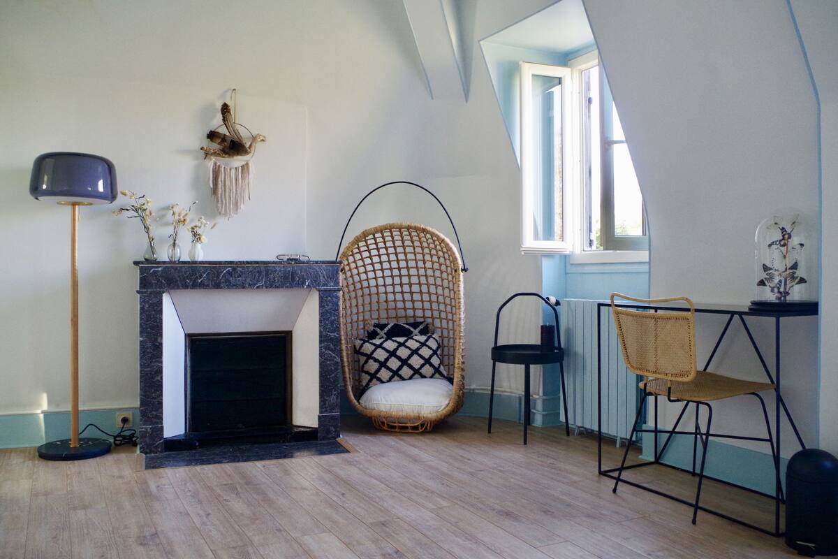 Um quarto com lareira, uma cadeira suspensa, uma mesa com cadeiras e uma janela com vista.