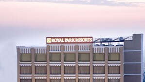 Um hotel com uma placa indicando "Royal Park Resorts" no topo. Possui vários andares com sacadas e uma área de estacionamento em frente.