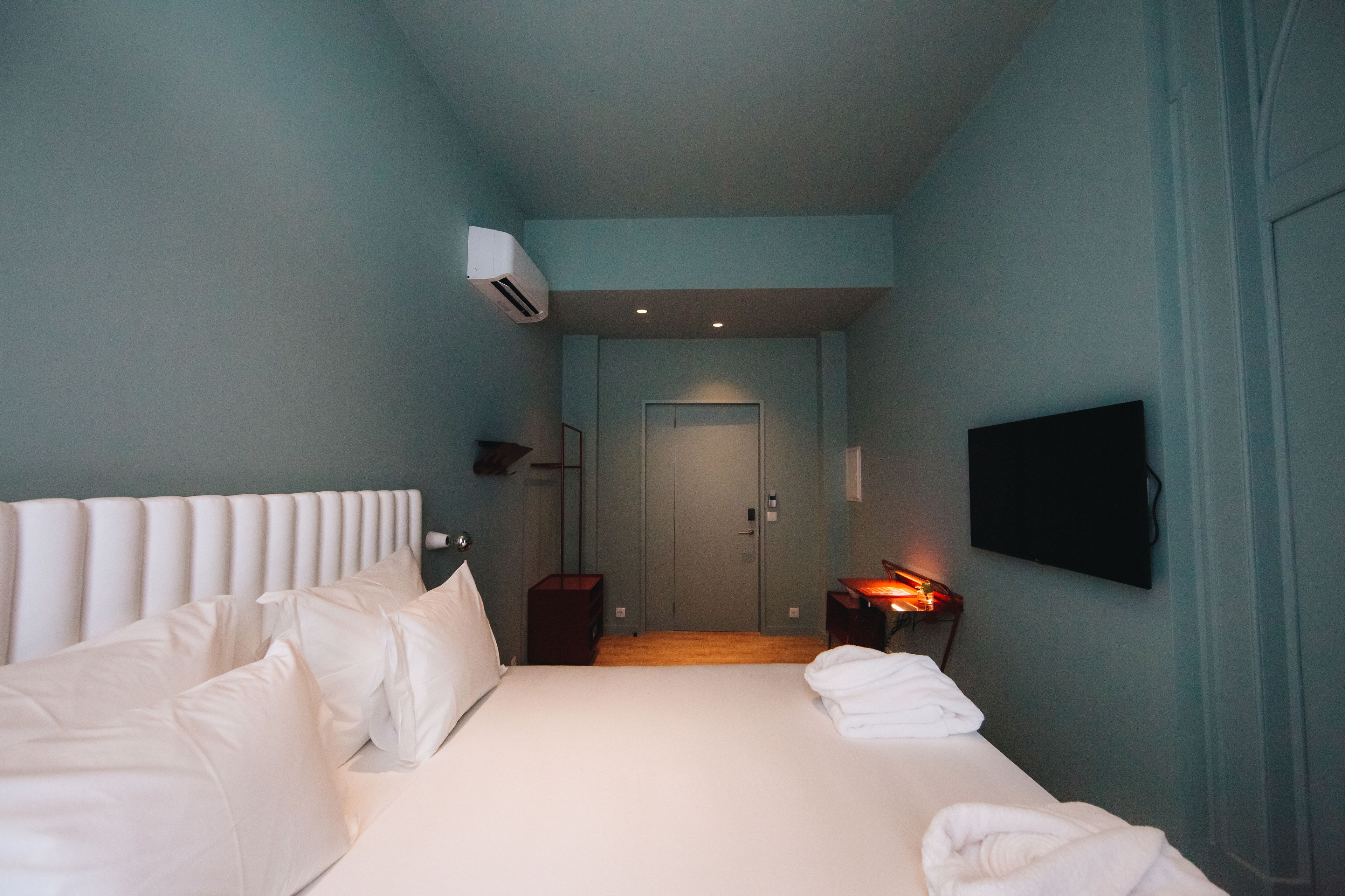 Quarto de hotel com cama, mesa de cabeceira, televisão fixada na parede e porta que dá para outro espaço.