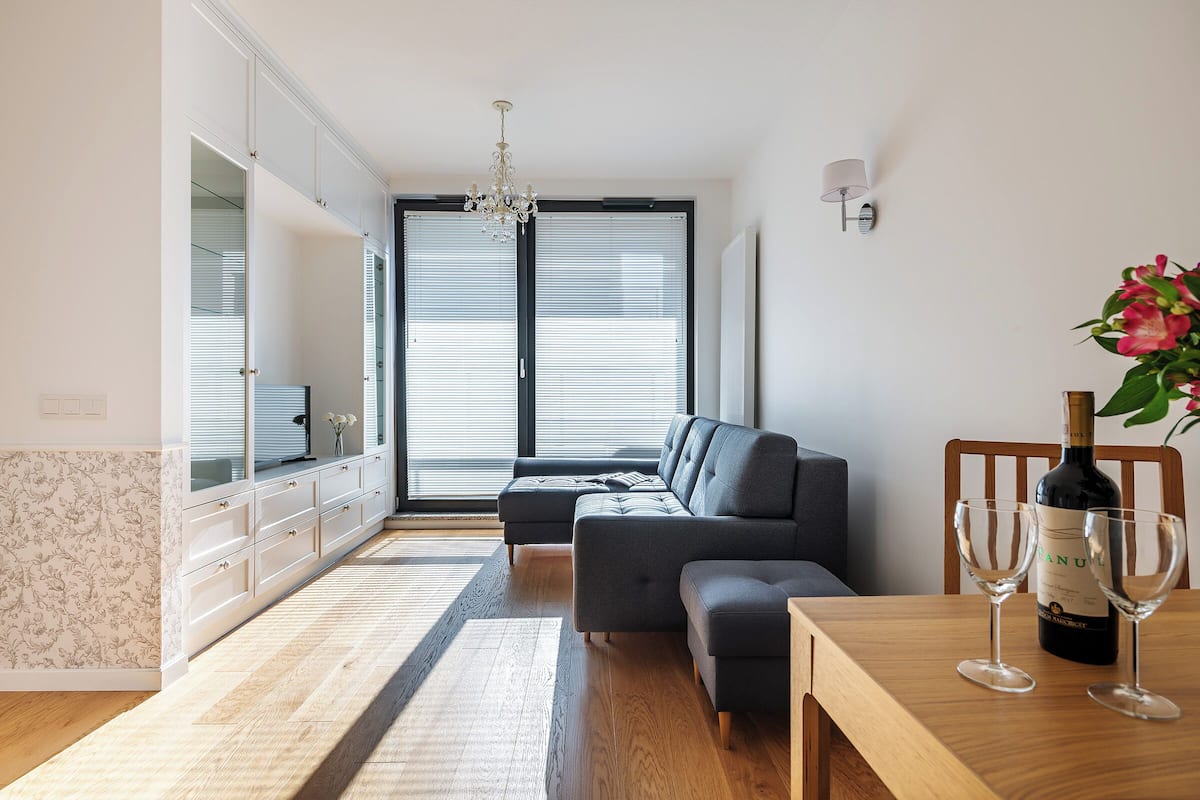 Uma sala de estar moderna com um sofá, uma mesa de jantar com copos de vinho e uma janela grande com persianas.