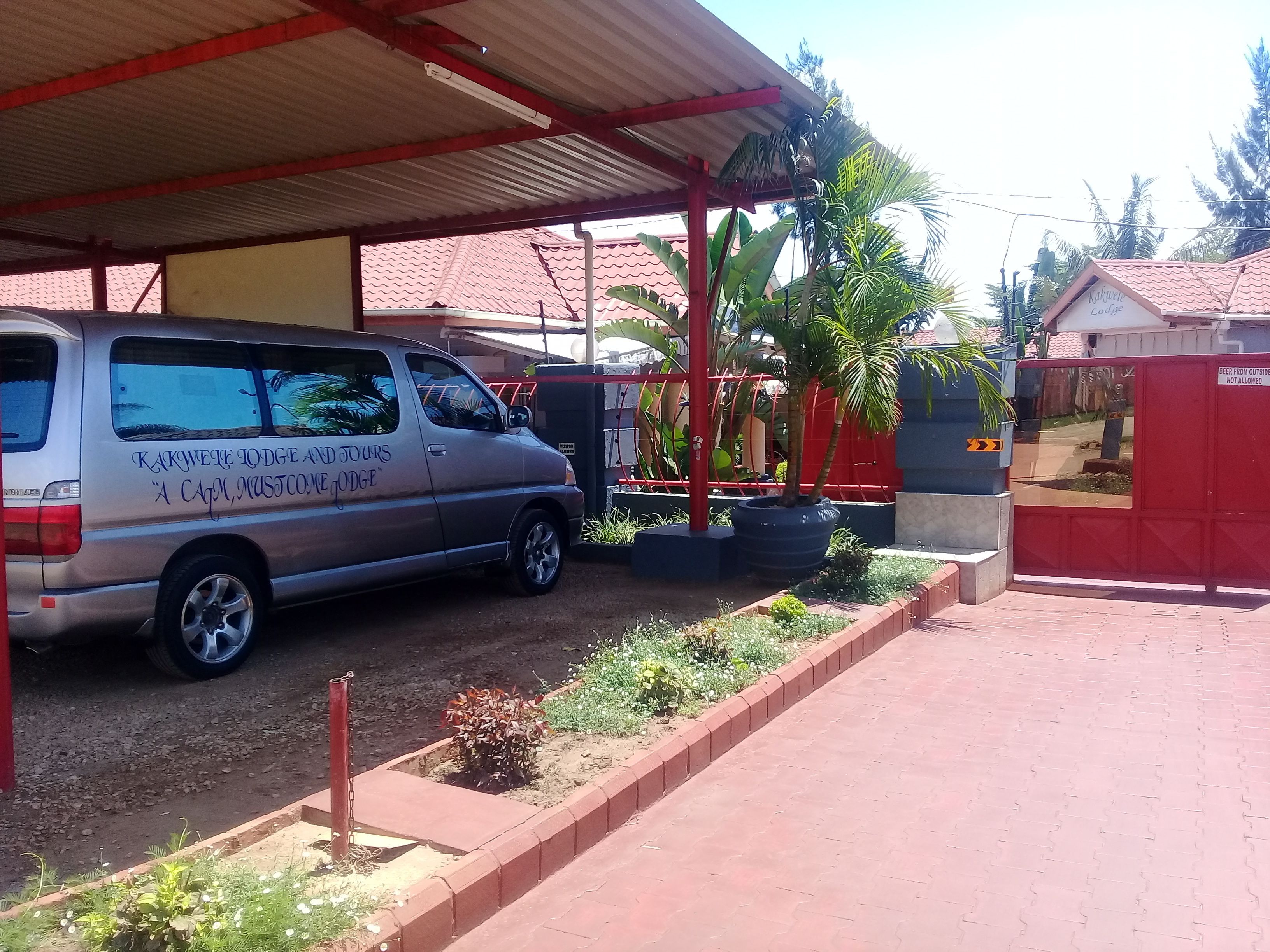 Uma van prateada estacionada sob uma cobertura, com uma placa indicando 'Kakwele Lodge and Tours'.