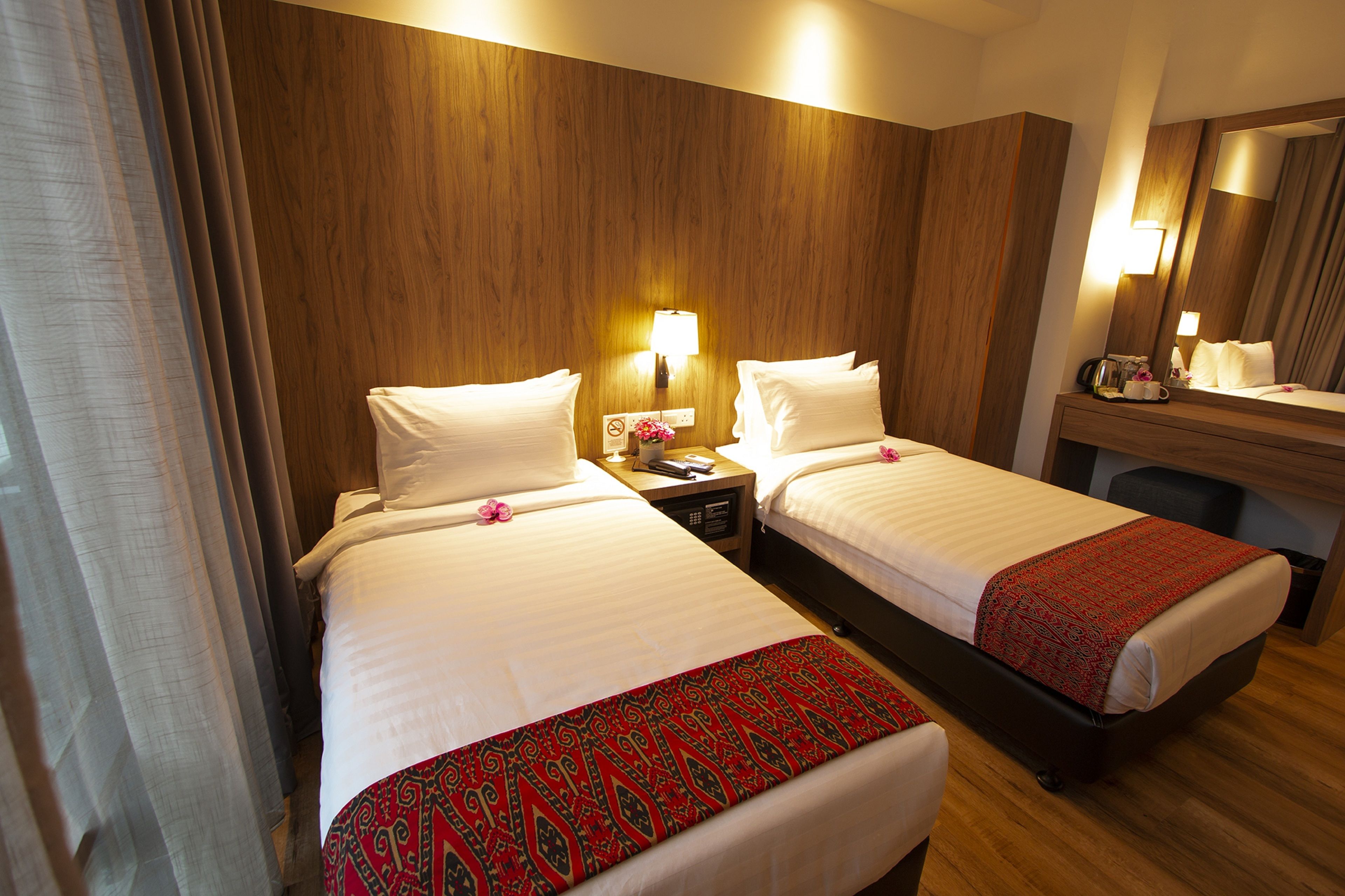 Quarto de hotel com duas camas, painéis de madeira nas paredes, uma escrivaninha com espelho e uma mesa de cabeceira com abajur e uma flor.