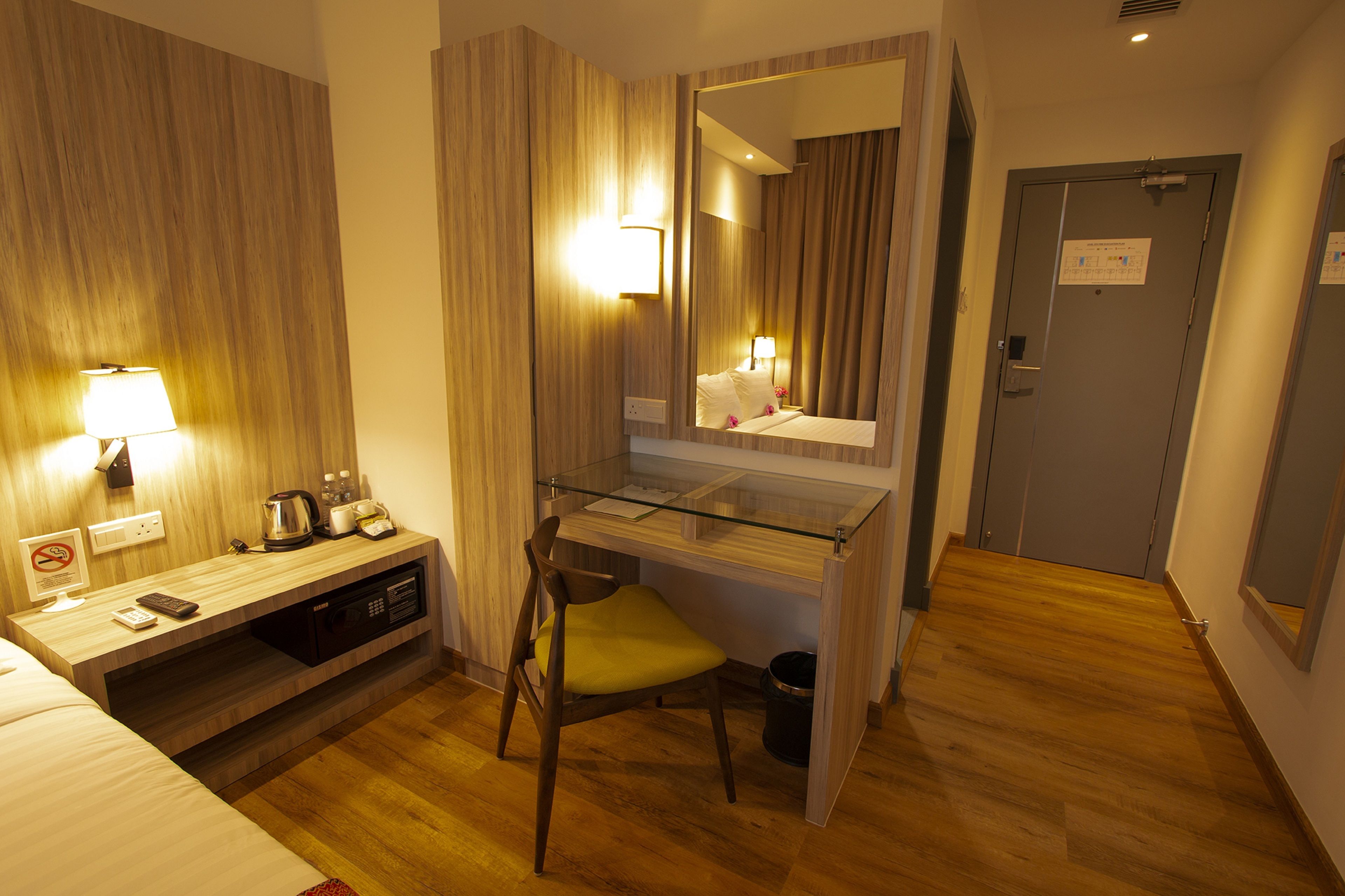 Quarto de hotel com piso de madeira, uma escrivaninha com cadeira, cama, espelho, abajur, telefone, chaleira e lixeira.
