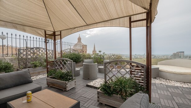 Um terraço no telhado com mobiliário externo, uma pérgula e vista para um edifício histórico.
