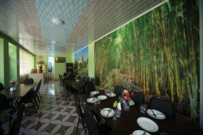 Uma sala de jantar com chão quadriculado, paredes verdes e um mural de floresta de bambu. Mesas dispostas com pratos, talheres e copos.