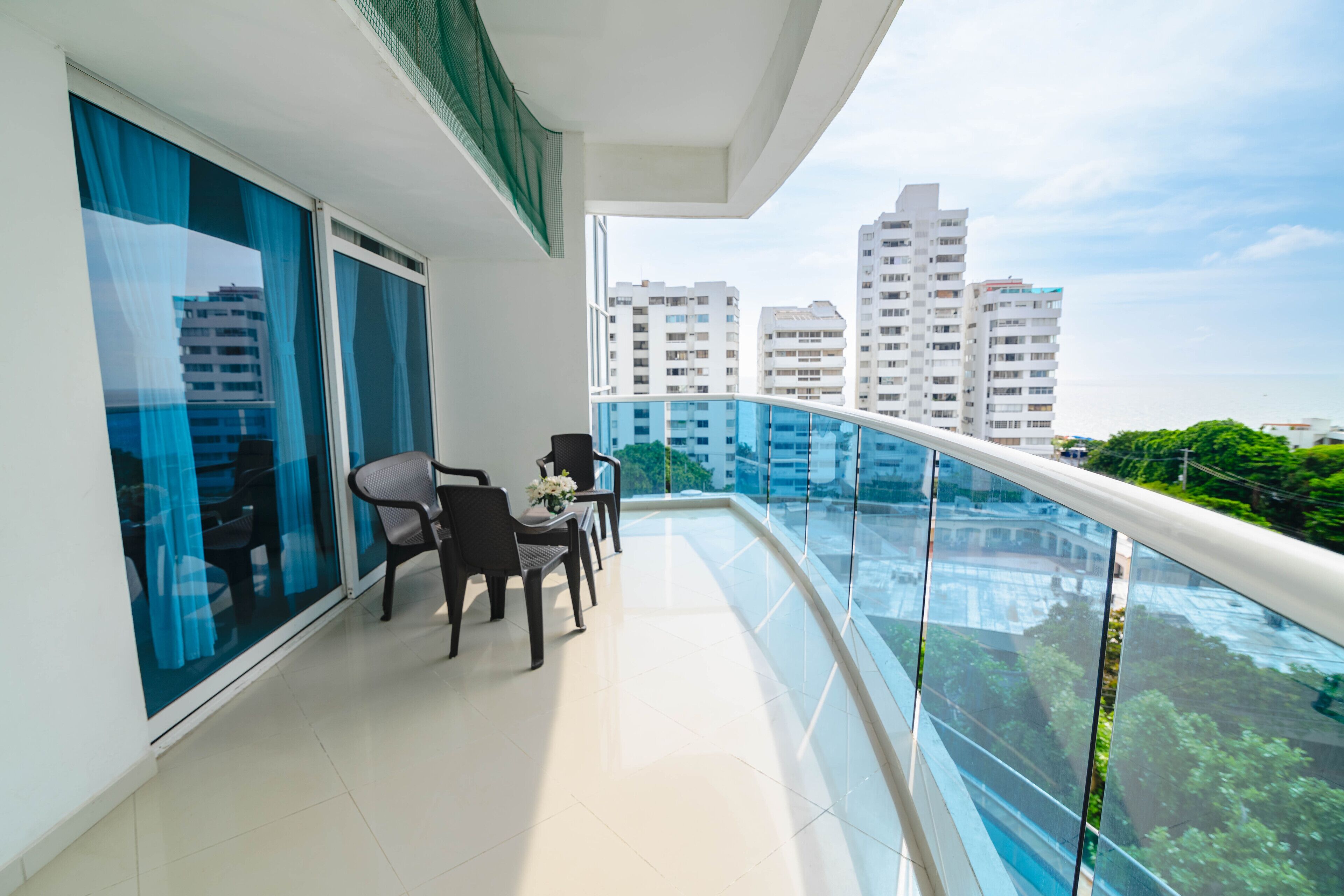 Uma varanda com guarda-corpo de vidro, uma mesa com cadeiras e vista para edifícios altos e áreas verdes.