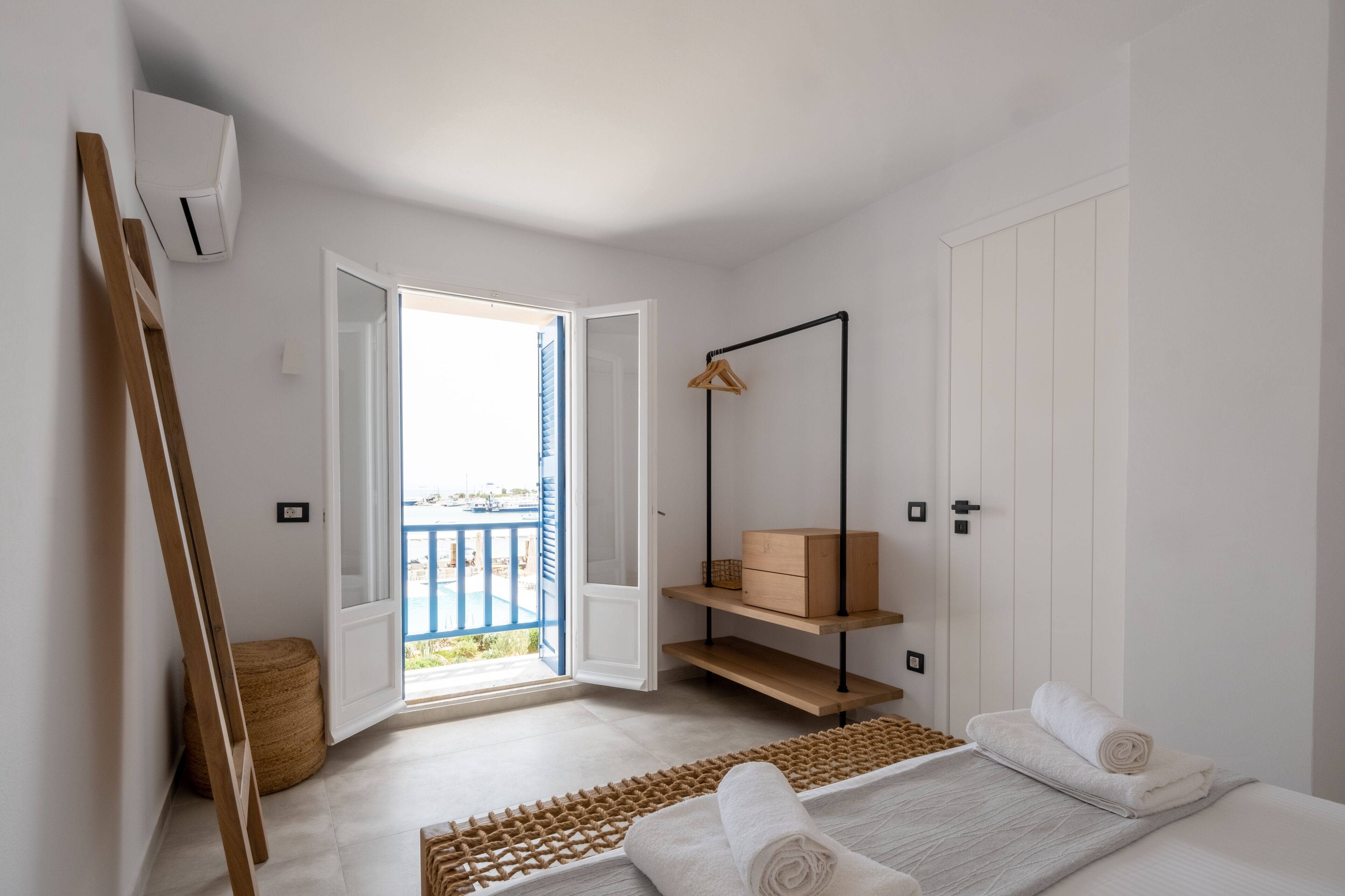 Quarto com cama, luminária pendente, prateleira de madeira, porta e vista para o exterior.