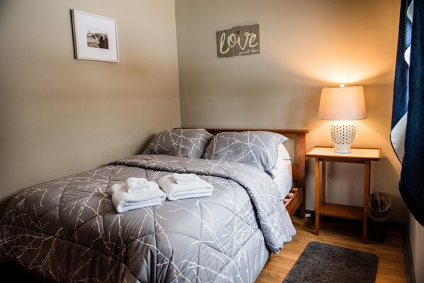 Uma cama bem arrumada com um edredom cinza e branco estampado, uma pequena mesa de cabeceira com uma luminária e um quadro na parede.