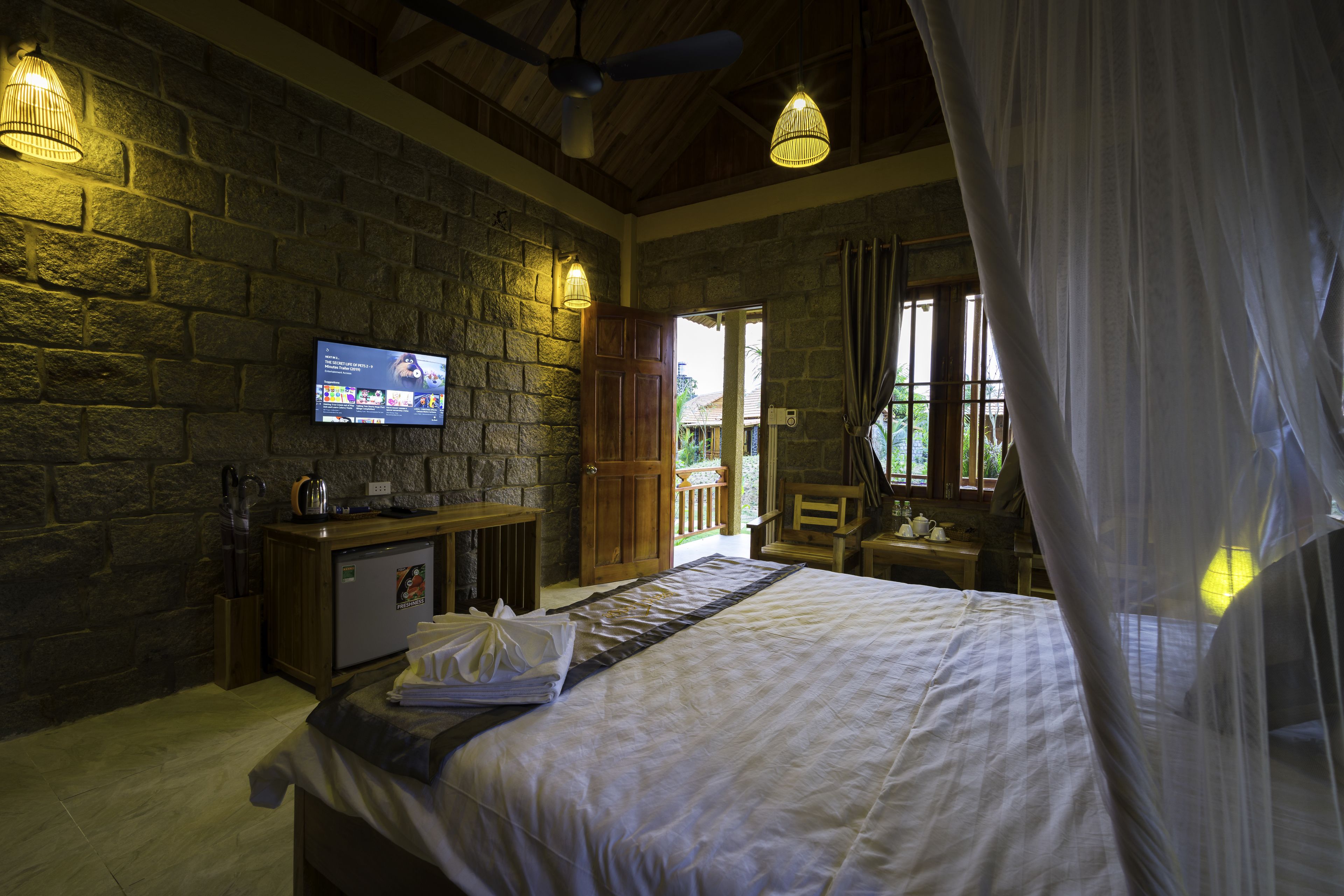 Quarto com parede de pedra, cama com roupa de cama branca, televisão na parede e escrivaninha de madeira com um computador.