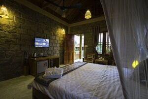 Quarto com parede de pedra, cama com roupa de cama branca, televisão na parede e escrivaninha de madeira com um computador.