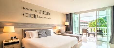 Quarto de hotel com uma cama grande, duas mesas de cabeceira com luminárias, um sofá e varanda com vista para uma casa e vegetação.