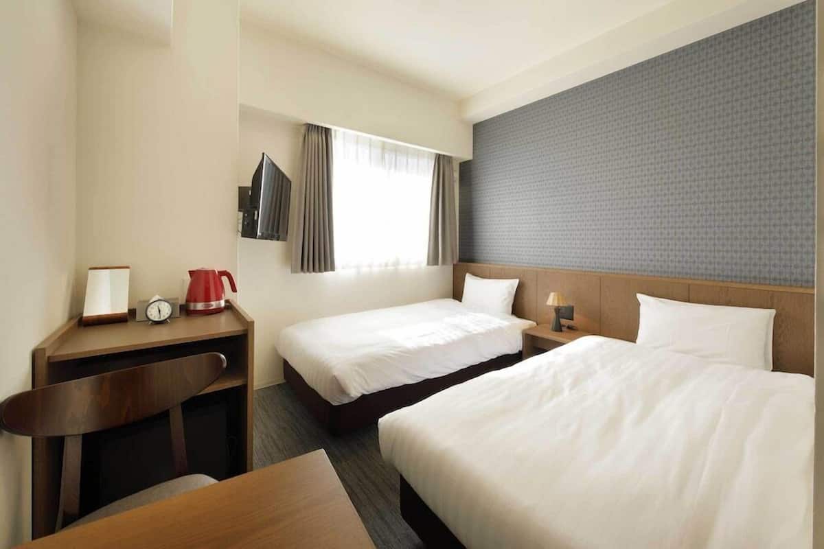 Quarto de hotel com duas camas, uma escrivaninha, uma televisão e uma janela com cortinas.