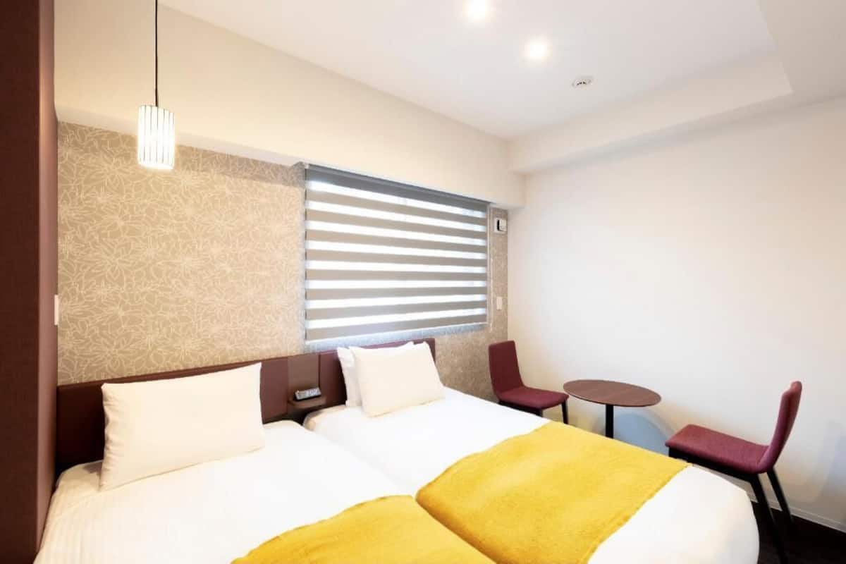 Quarto de hotel com uma cama grande, uma mesinha, duas cadeiras, uma janela com persianas e parede com padrão.