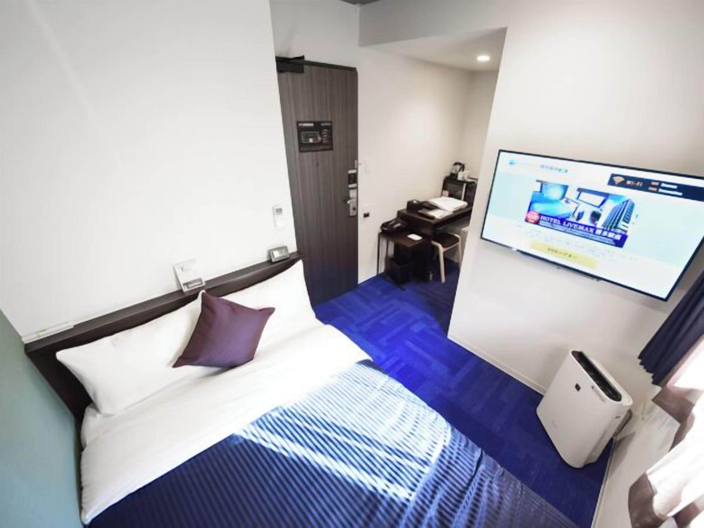 Quarto de hotel com cama, escrivaninha, cadeira, computador e televisão exibindo um anúncio do hotel.