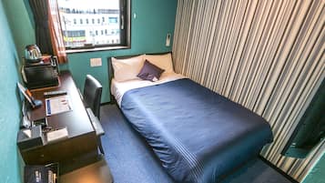 Quarto de hotel com cama, escrivaninha, cadeira, mala e janela com vista para edifícios.