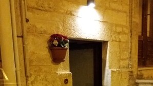 Um edifício de pedra com uma pequena entrada, um vaso de flores na parede e um candeeiro sobre a porta.