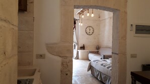 Quarto rústico com parede de pedra, cama com colcha estampada, mesa de cabeceira de madeira, relógio de parede e luminária suspensa.
