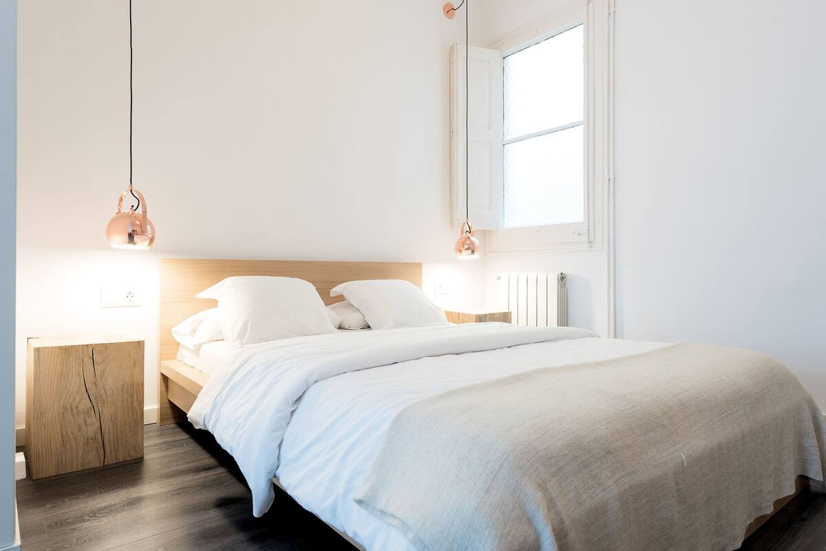 Quarto minimalista com cabeceira de madeira, cama grande com roupa de cama branca e luminárias pendentes.