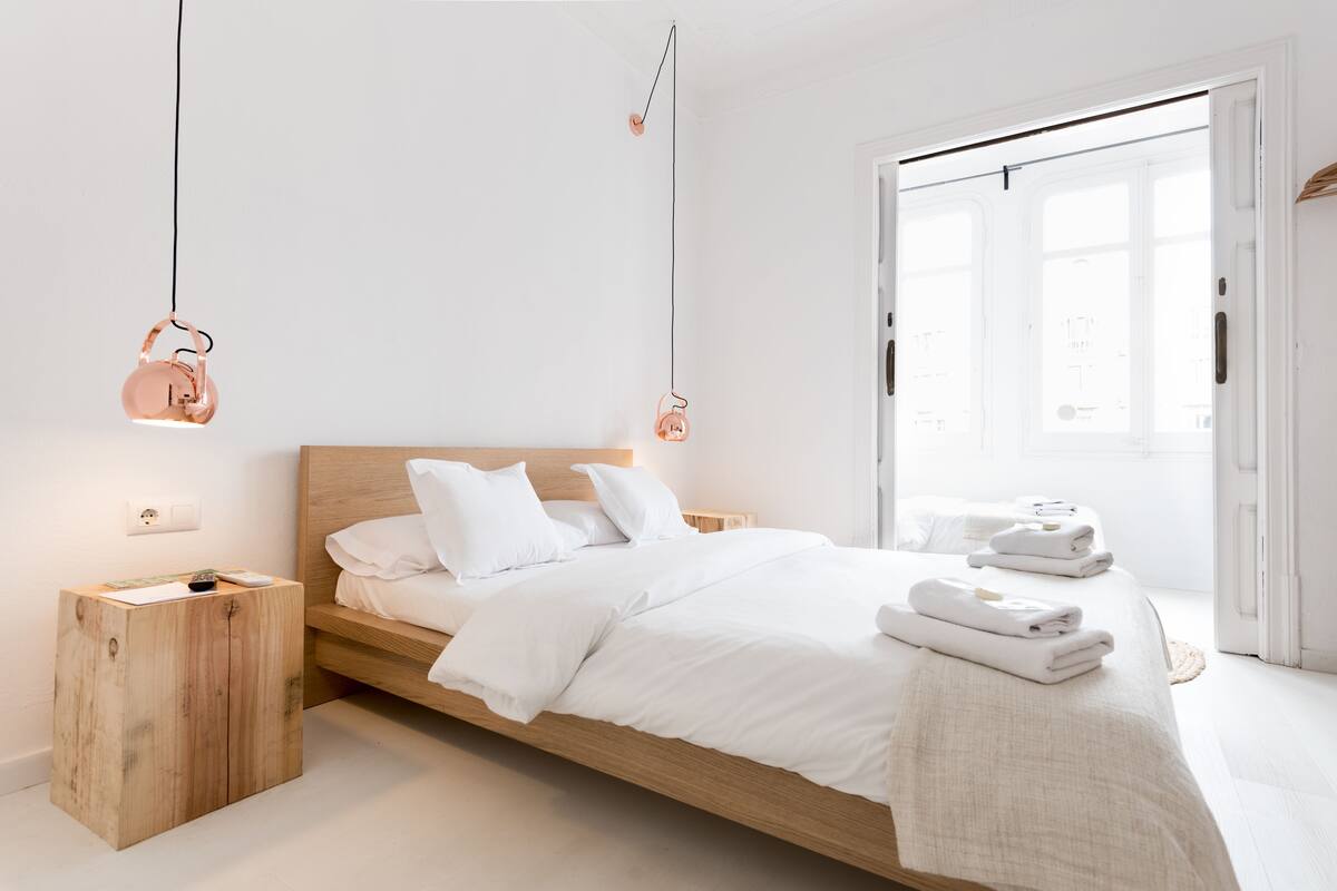 Quarto minimalista com cama de madeira, roupa de cama branca e mesa de cabeceira com abajur.