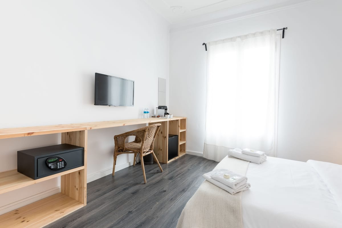 Quarto de hotel com escrivaninha de madeira, uma cadeira, TV fixada na parede, cama com roupa de cama branca e janela com cortinas.