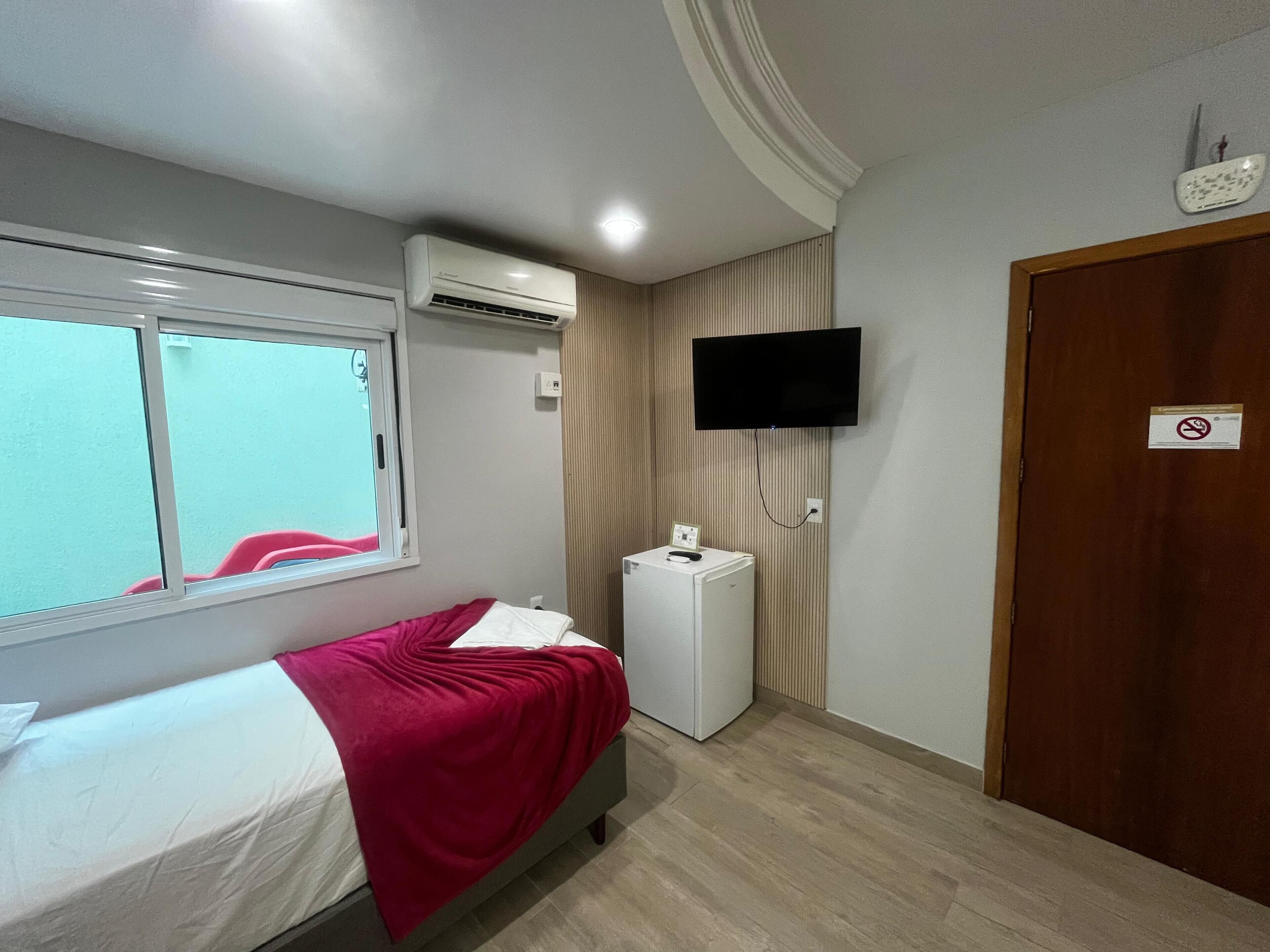 Quarto de hotel com uma cama, televisão na parede, janela, ar condicionado e porta com placa de proibido fumar.