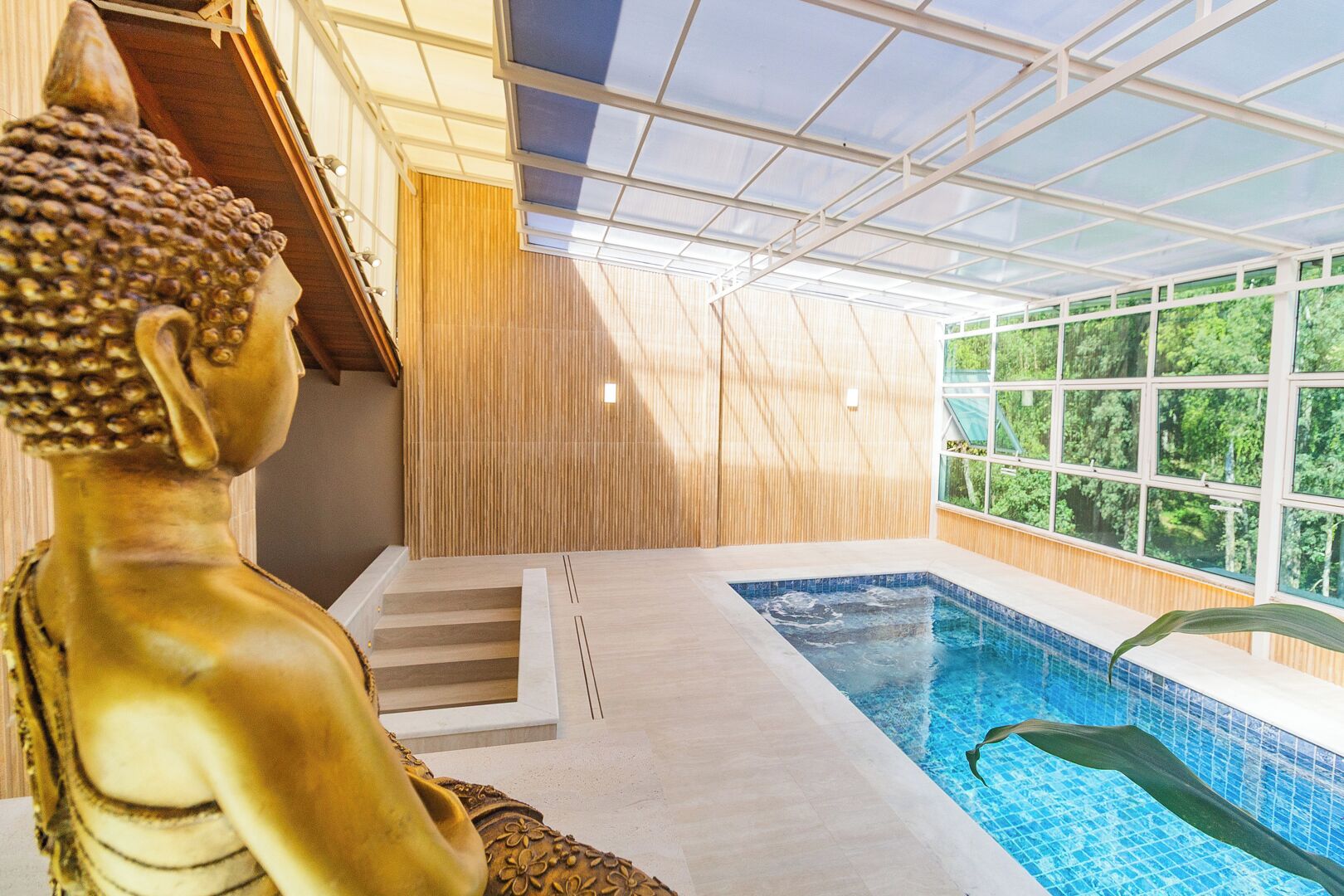 Uma estátua dourada de Buda, uma piscina e vista para o verde através de grandes janelas.