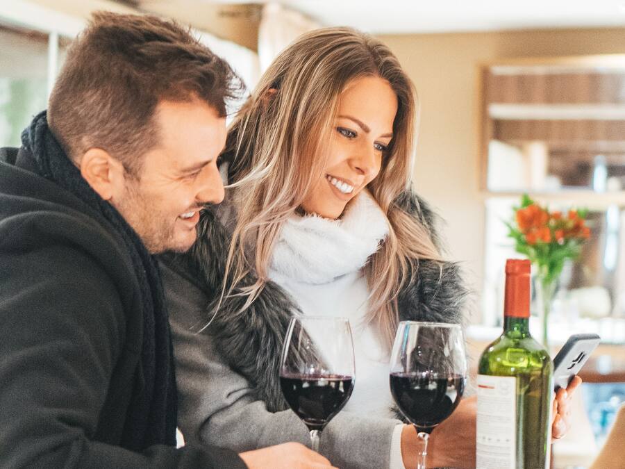 Um casal sentado em uma mesa redonda com duas taças de vinho e uma garrafa de vinho.