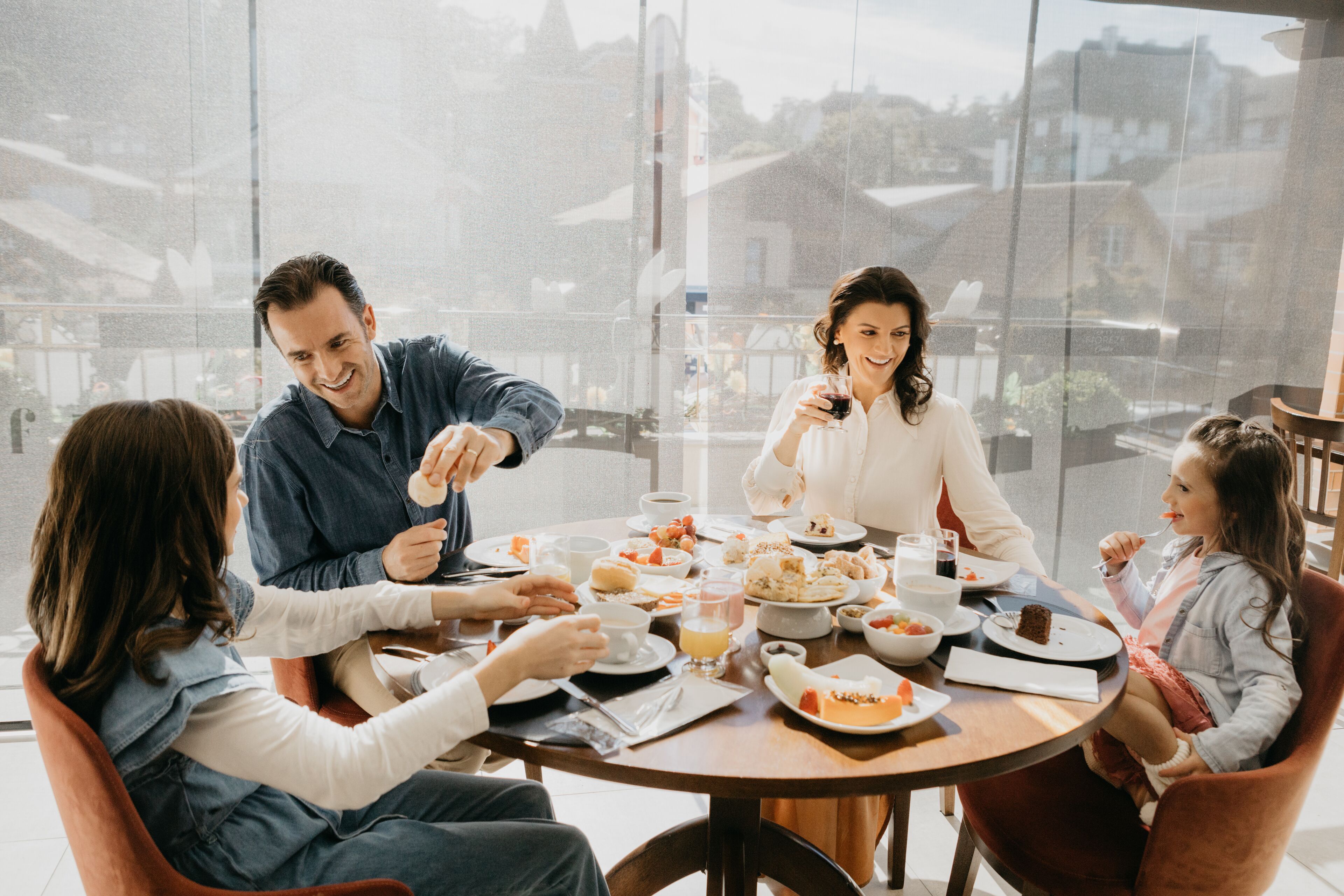 Uma família jantando à mesa com vista para a cidade, incluindo um copo de suco e diversos pratos.
