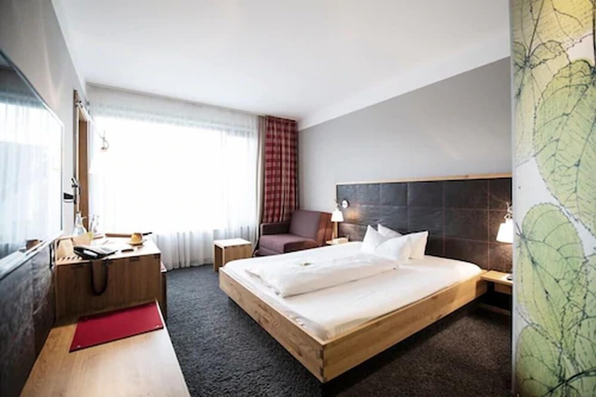 Quarto de hotel com cama, escrivaninha, cadeira, mesa de cabeceira e janela com cortinas.