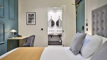Quarto de hotel com cama, escrivaninha com luminária, cadeira, banheiro com pia e espelho, e porta com puxador.