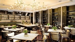 Um restaurante com várias mesas e cadeiras, um lustre moderno e prateleiras com objetos nas paredes.
