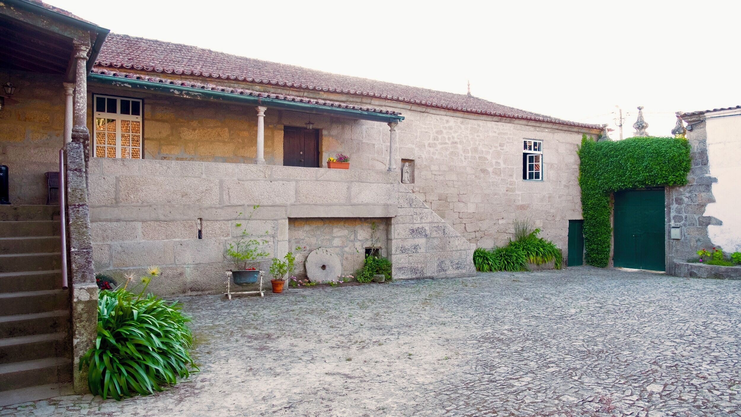 Um edifício de pedra com telhado ladrilhado, uma pequena escadaria e um pátio de calçada.
