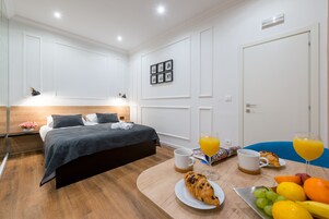 Quarto de hotel moderno com cama bem arrumada, mesa de pequeno-almoço com croissants, fruta e sumo, e parede com quadros emoldurados.