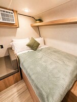 Uma cama compacta com um cobertor verde, uma cabeceira de madeira, uma pequena prateleira com uma planta e um ar condicionado fixado na parede.